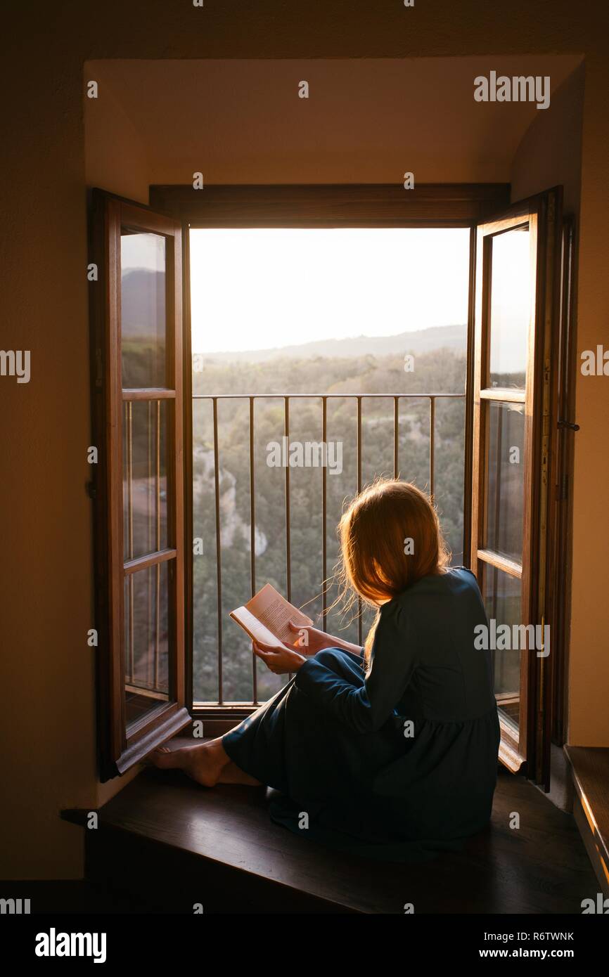 girl is sitting on the floor by the open window and reading a book ...