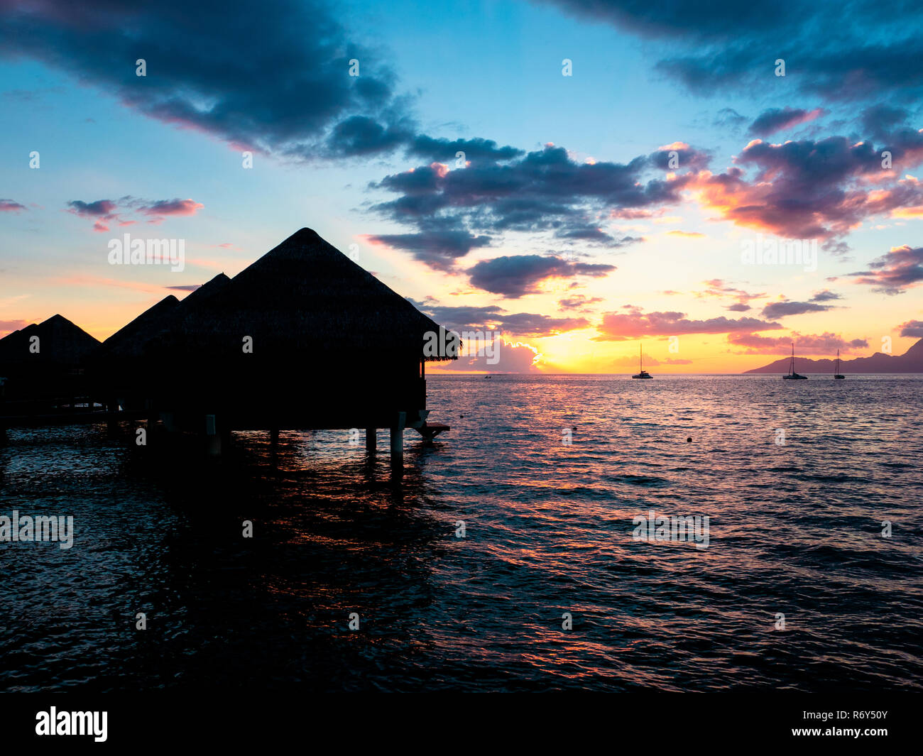 Sunset Over Thatched Roof Overwater Cabanas with Sailboats in Tahiti ...