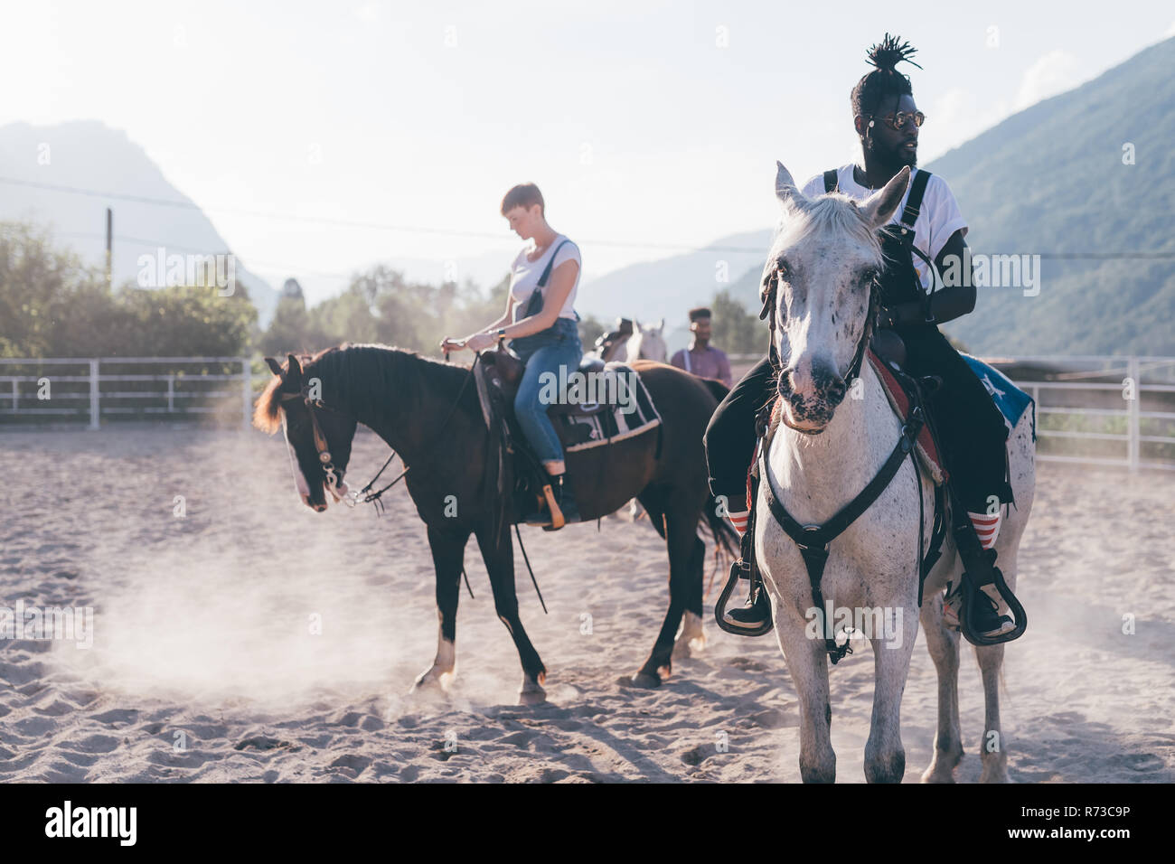 Young hipster man and woman riding horses in rural equestrian arena ...