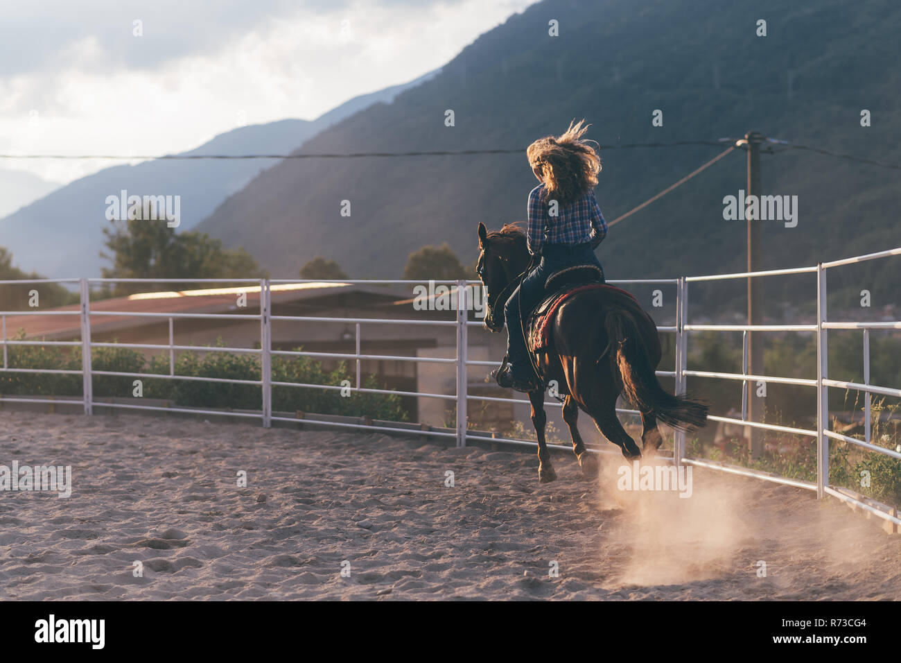 Long haired young woman cantering on horse in rural equestrian arena ...