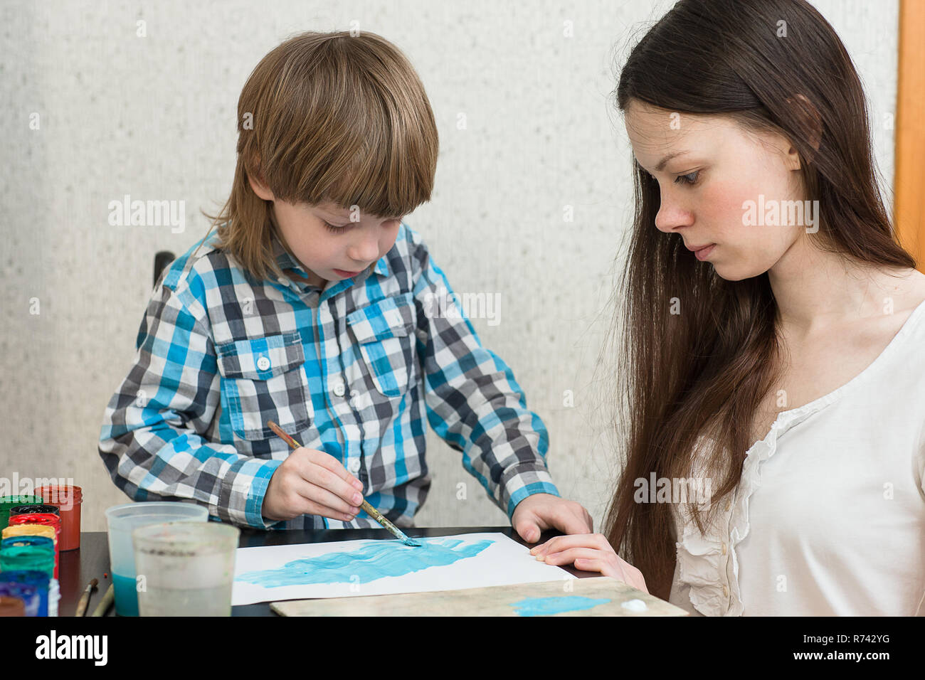 Young beautiful mother with boys son paint colors at home. Mom teaches ...