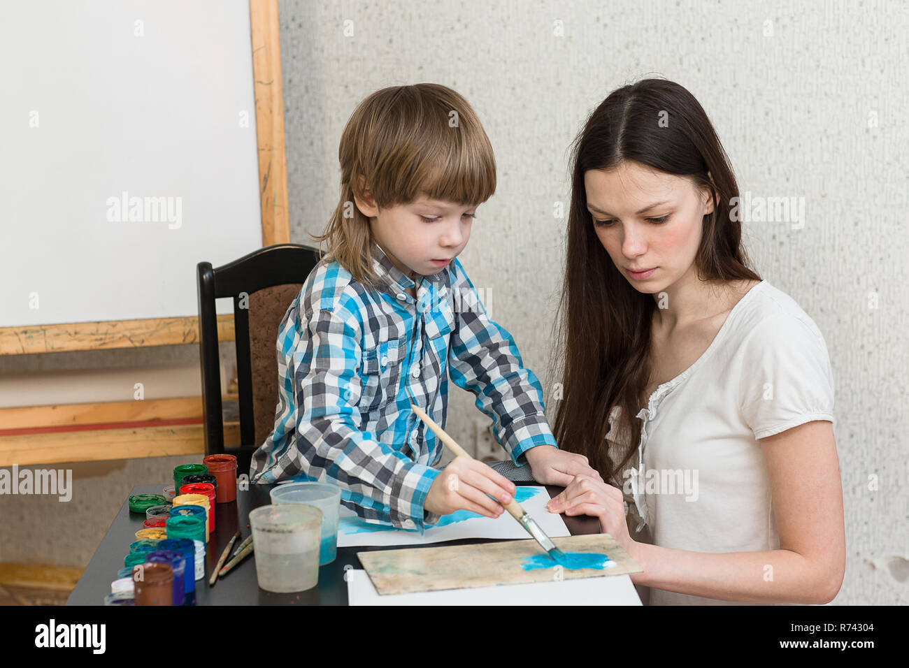 Young beautiful mother with boys son paint colors at home. Mom teaches ...