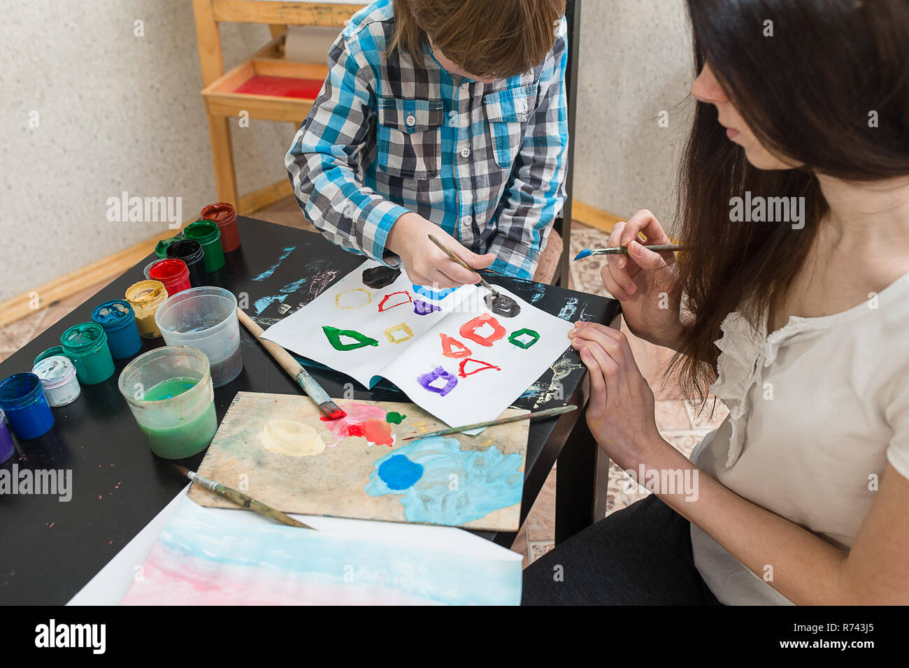 Young beautiful mother with boys son paint colors at home. Mom teaches ...