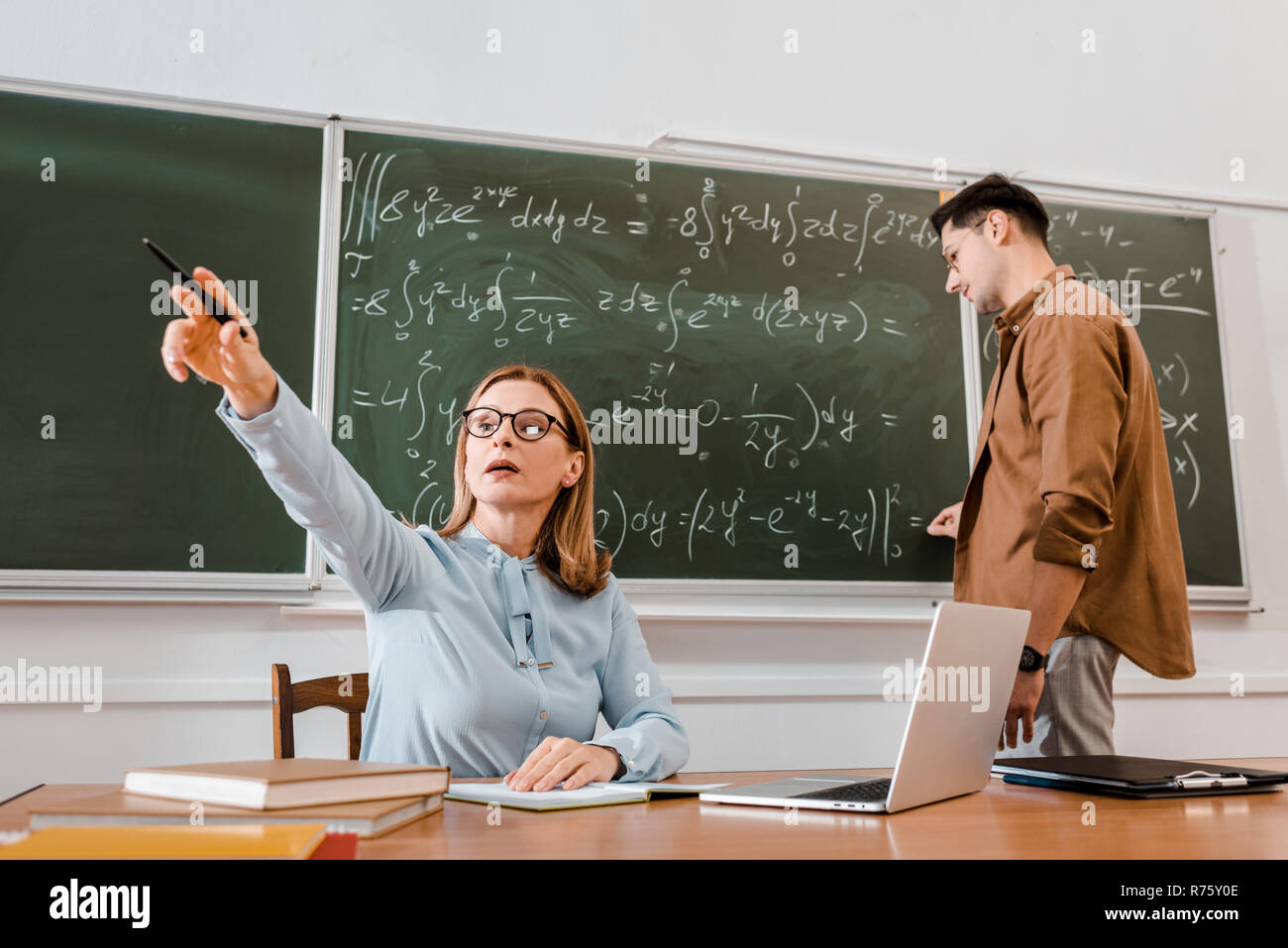 Female teacher sitting at desk and pointing at something in classroom ...