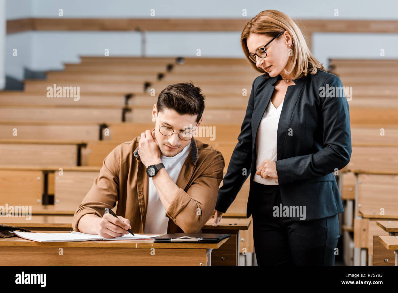 female university teacher looking at male student writing exam in ...
