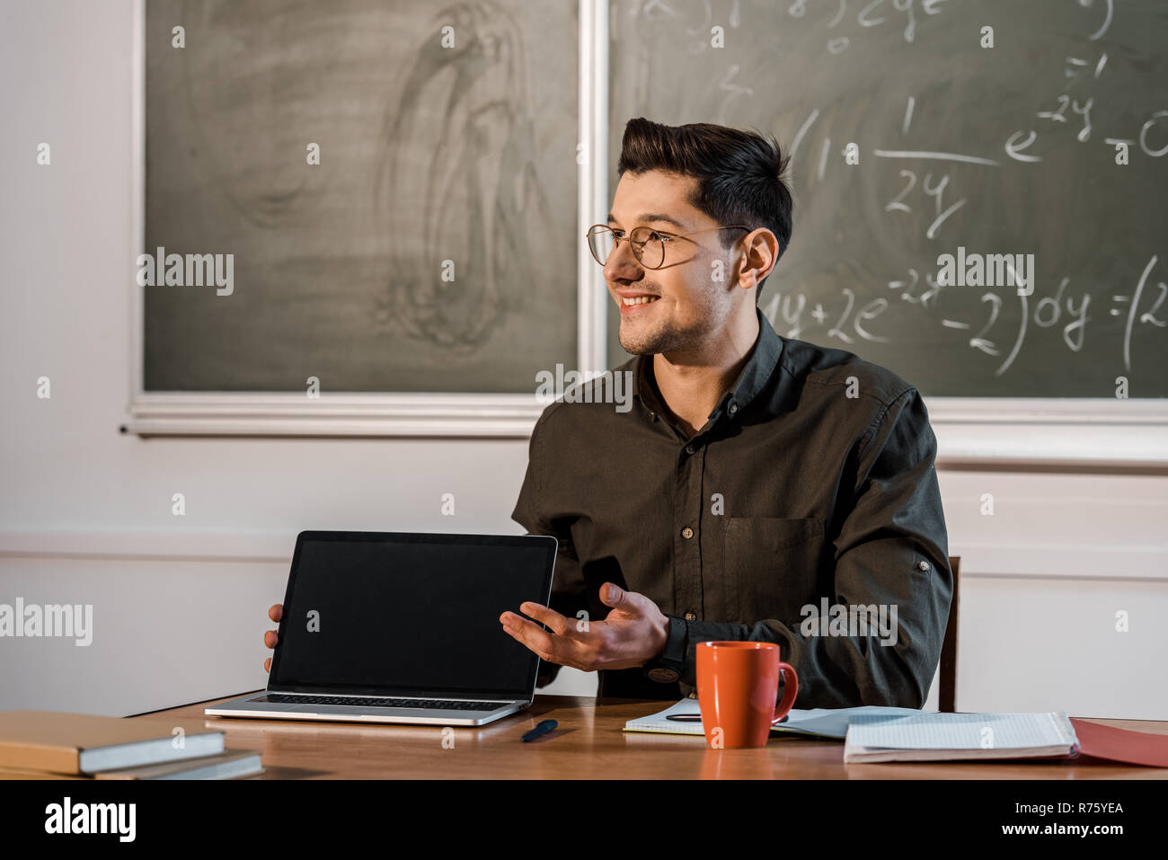 smiling male teacher sitting at desk and showing laptop with blank ...