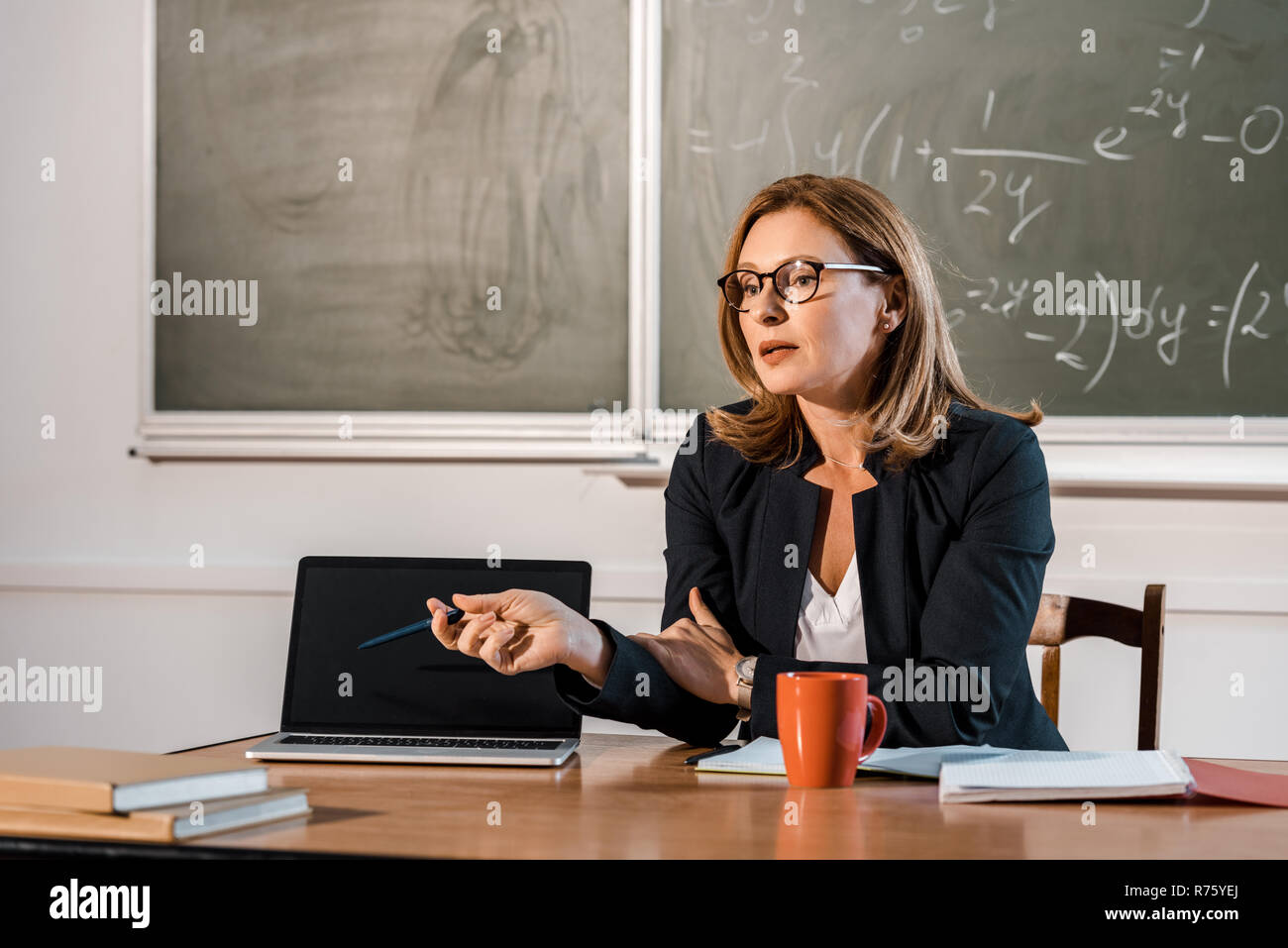 female teacher sitting at desk and pointing at laptop with blank screen ...
