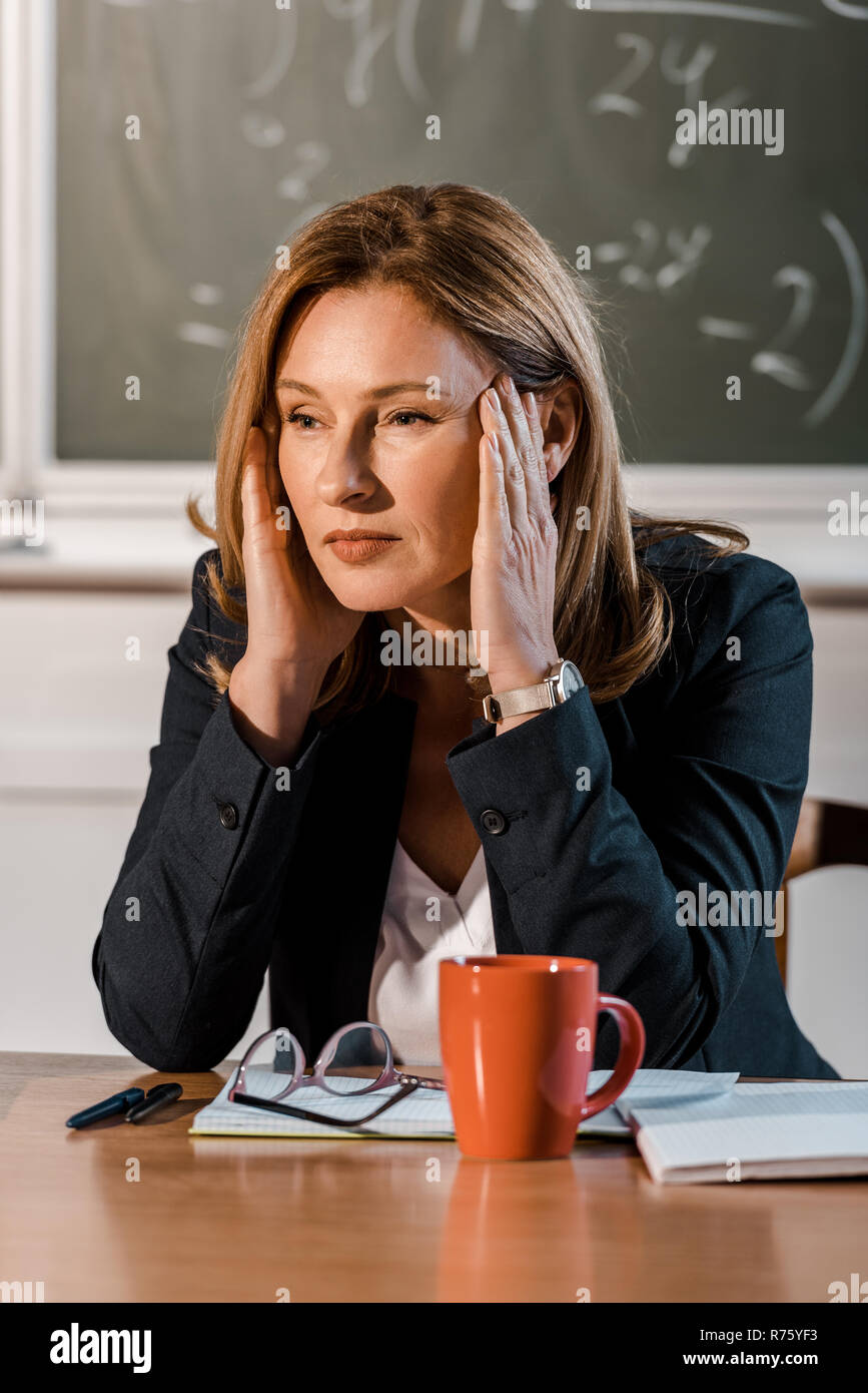 female teacher sitting at desk with hands on head and having headache ...