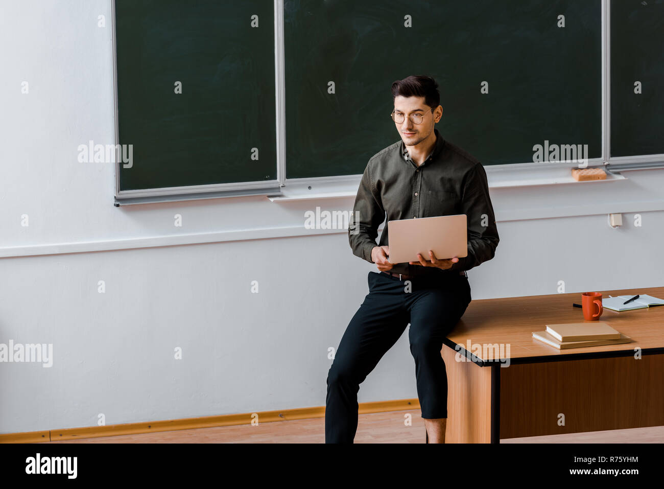 male teacher in formal wear sitting on desk and holding laptop in ...