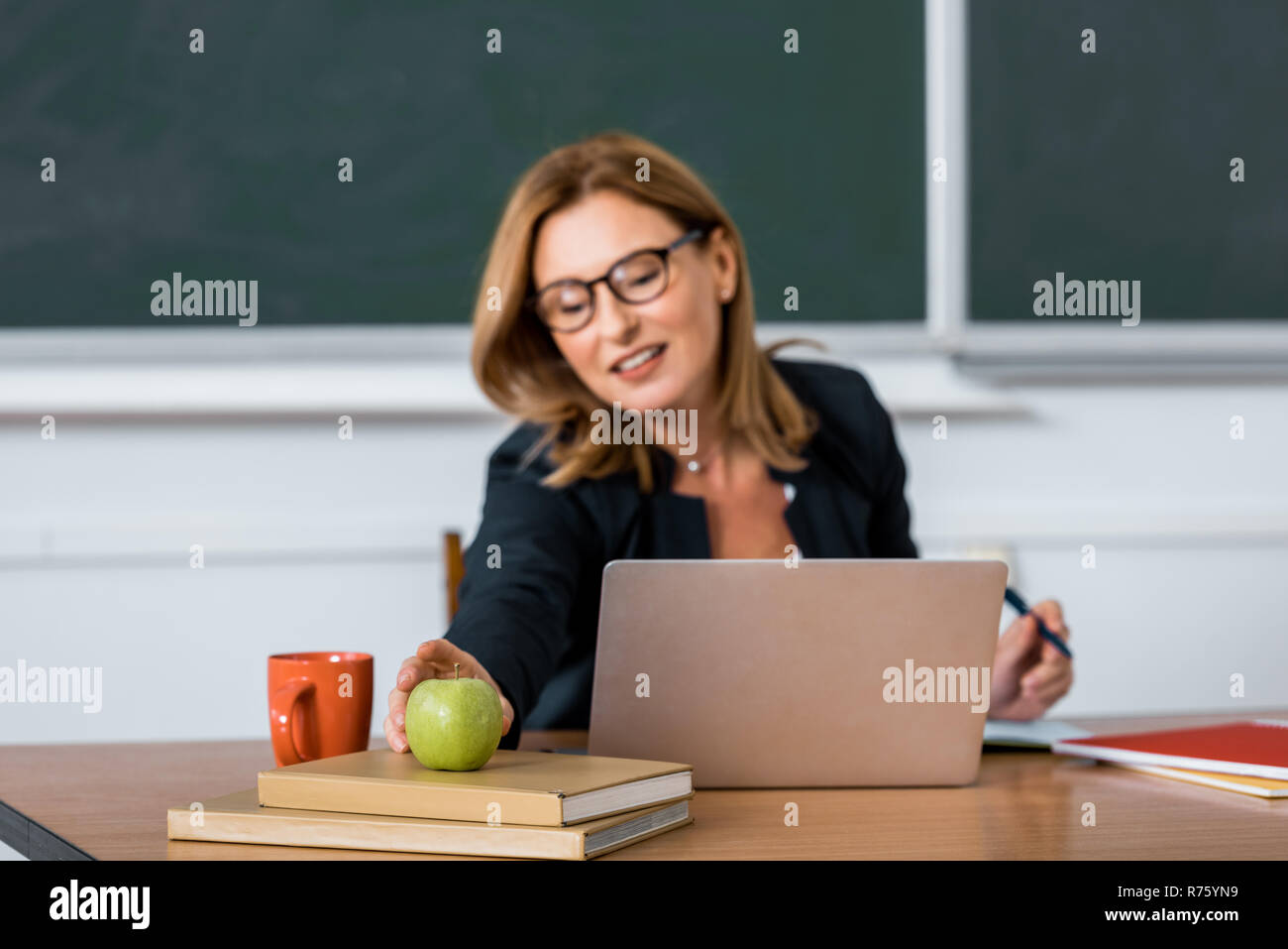 female teacher sitting at computer desk and reaching for apple in ...