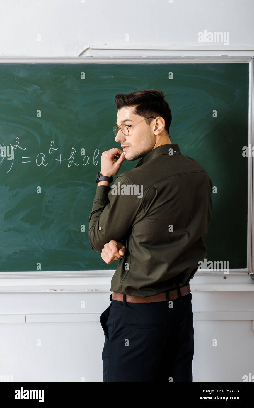 pensive male teacher touching chin and solving equation on chalkboard ...