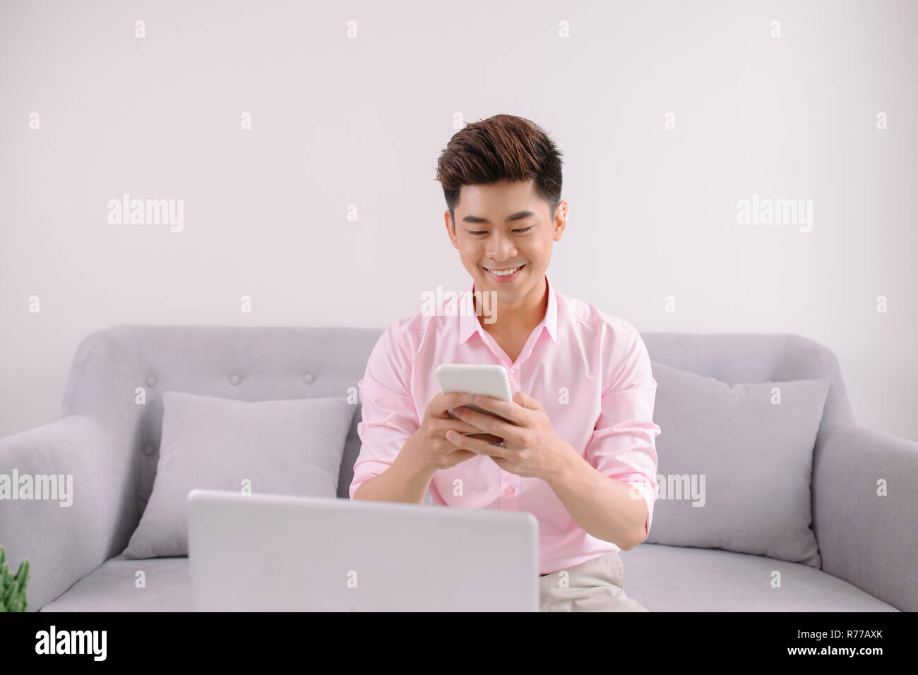 Young smiling asian man sitting on couch texting while working on ...