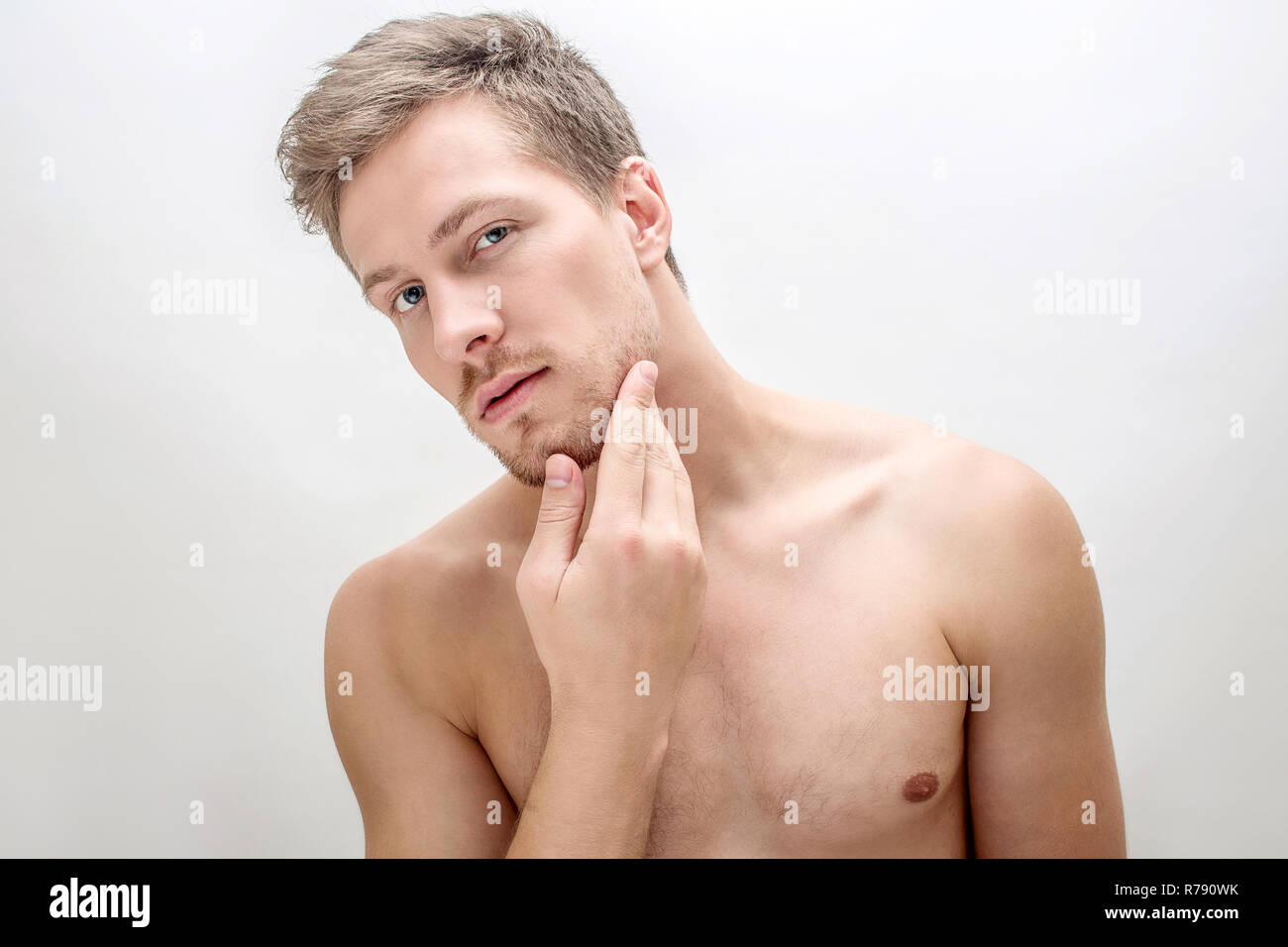 Handsome young man looking straight. He touches face. Guy is shirtless ...