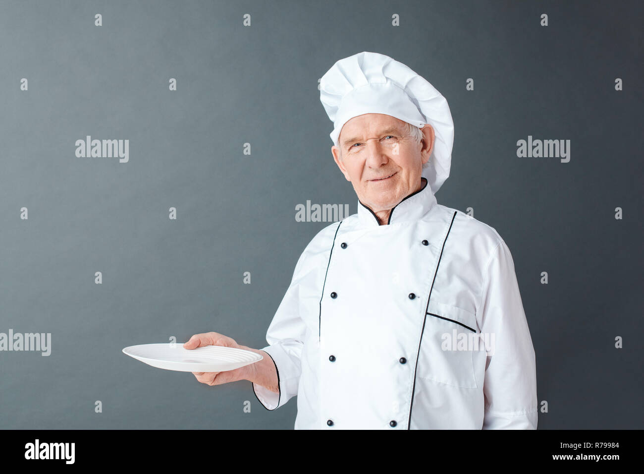 Senior chef studio standing isolated on gray with empty plate looking ...