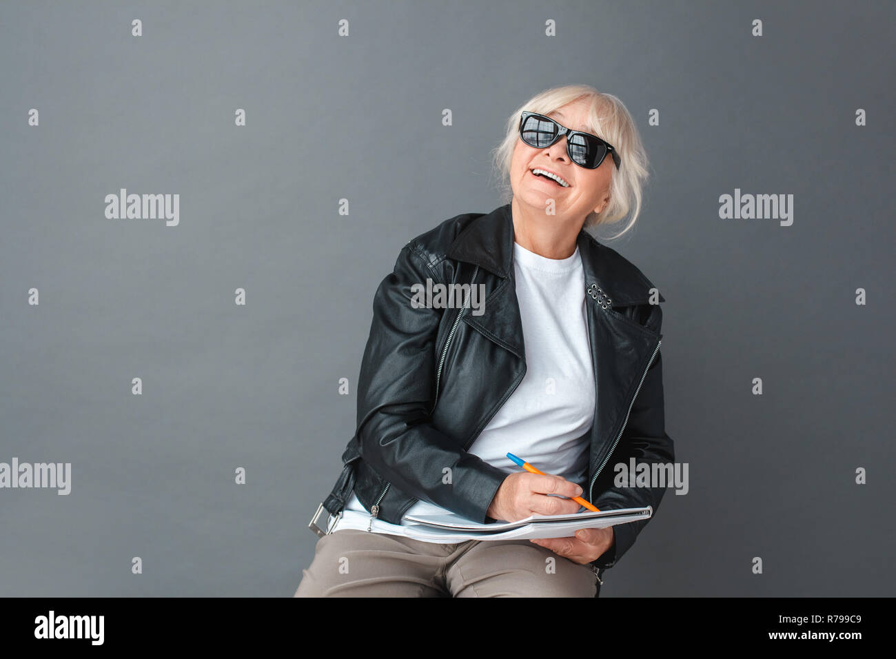 Senior woman in leather jacket and sunglasses studio sitting isolated ...