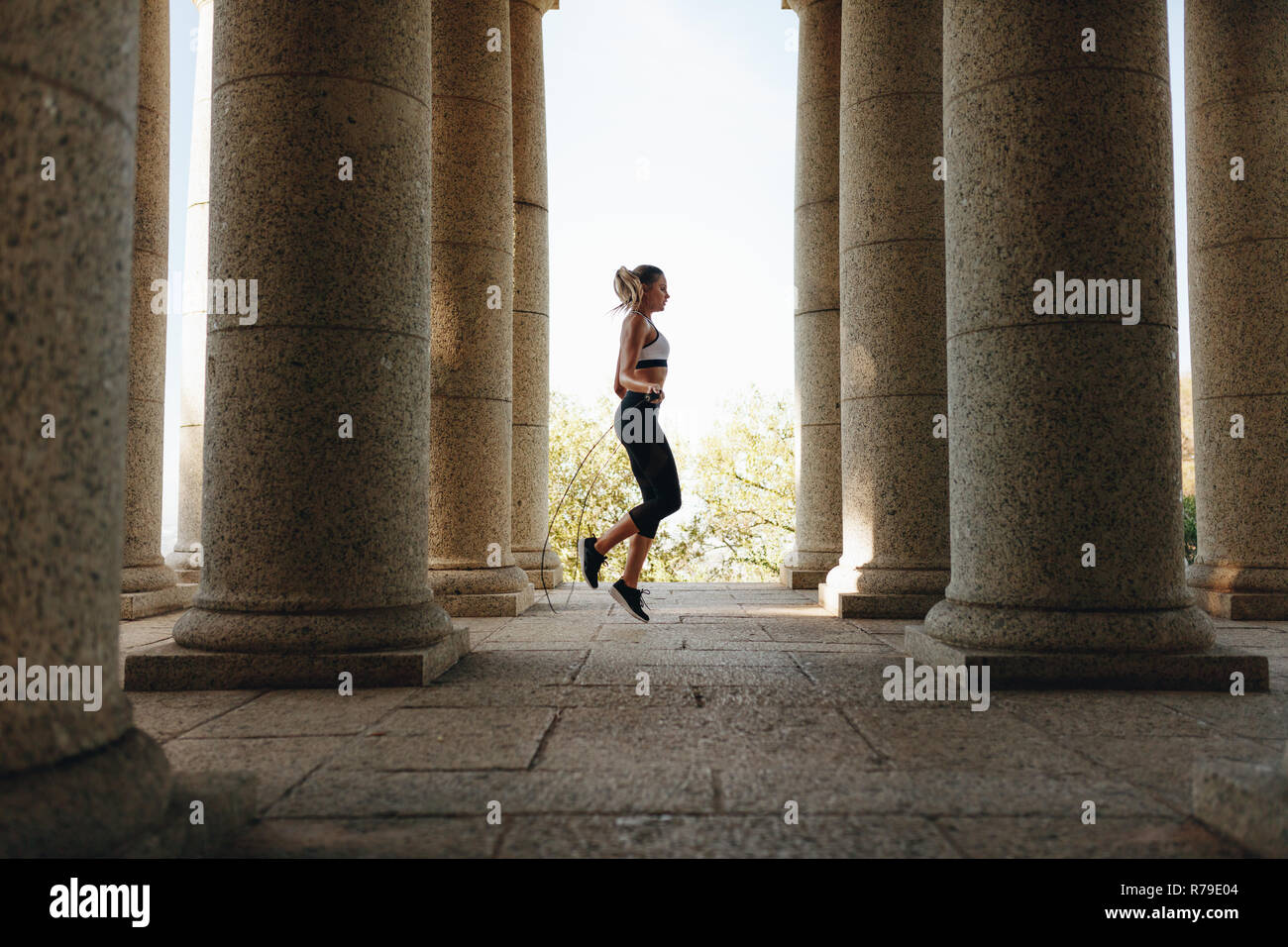 Woman in fitness wear doing skipping standing near stone pillars. Side ...