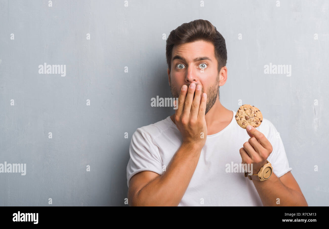 Handsome young man over grey grunge wall eating chocolate chip cooky ...