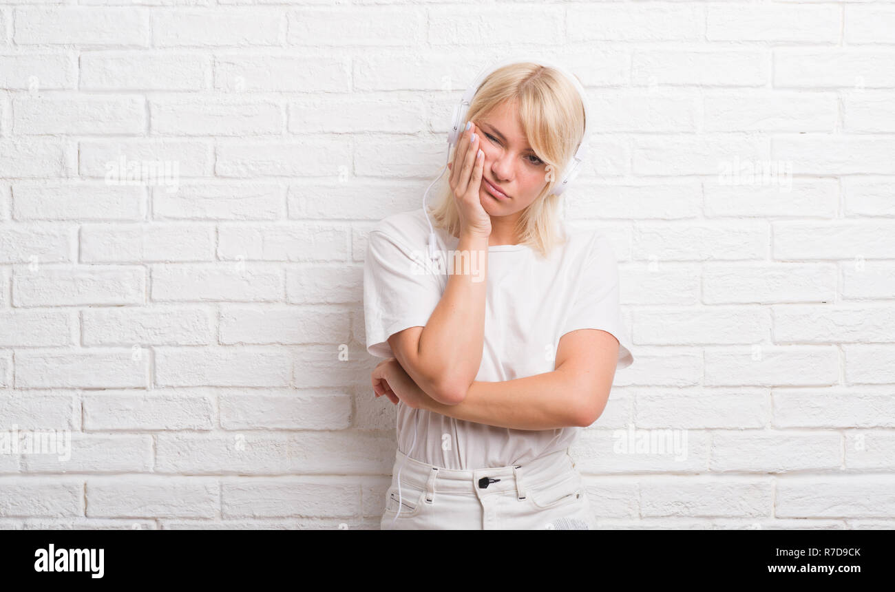 Adult caucasian woman over white brick wall wearing headphones thinking ...