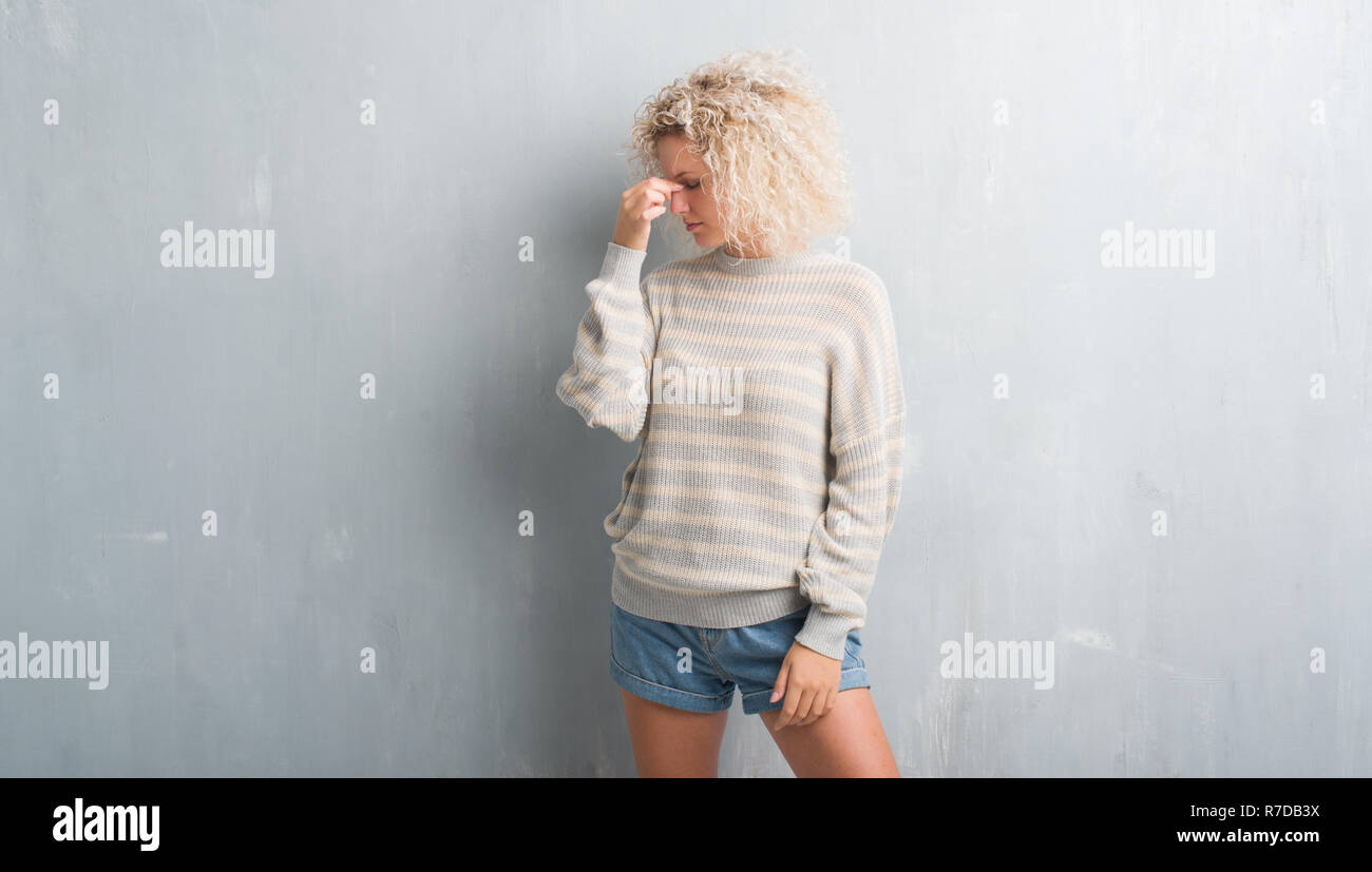 Young blonde woman with curly hair over grunge grey background tired ...