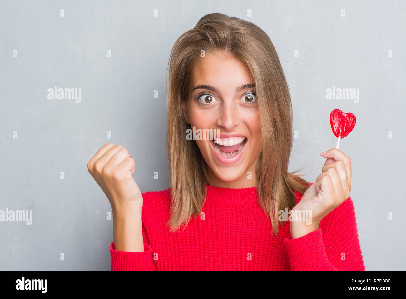 Beautiful young woman over grunge grey wall eating red heart lollipop ...