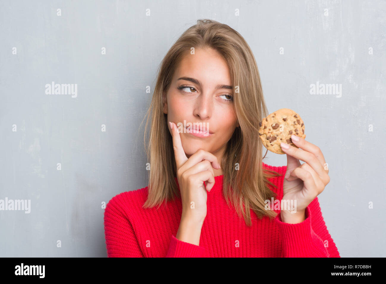 Beautiful young woman over grunge grey wall eating chocolate chip cooky ...