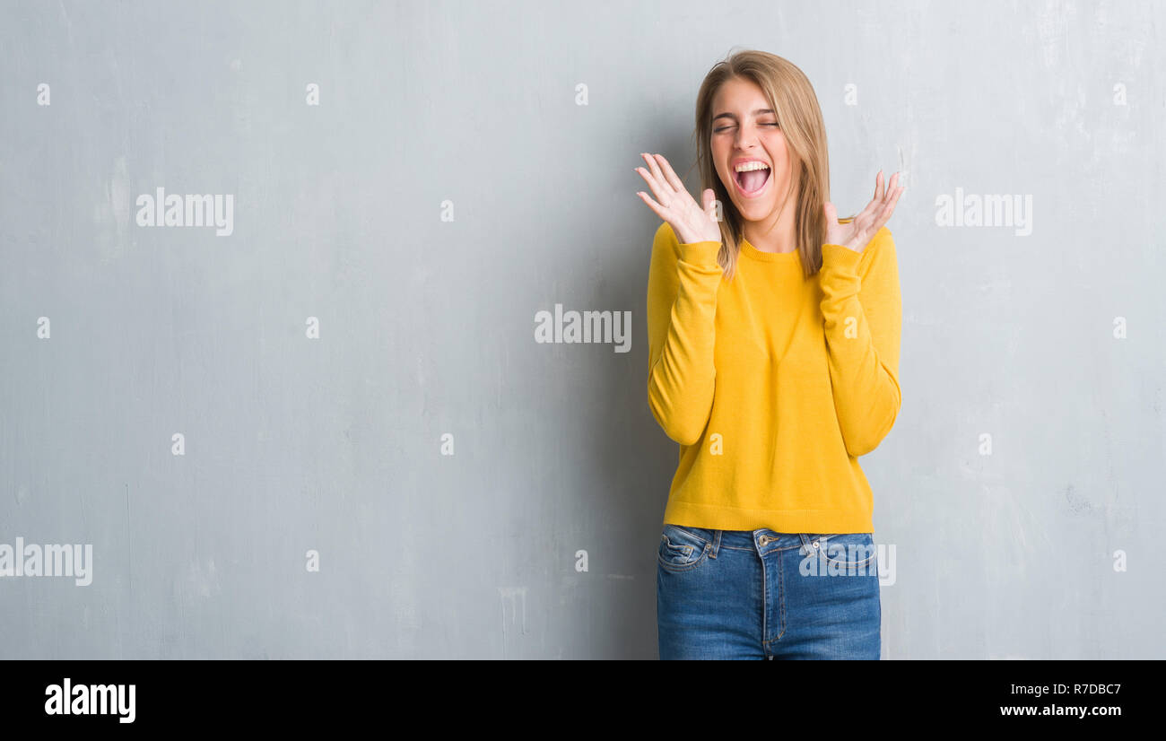 Beautiful young woman standing over grunge grey wall celebrating mad ...