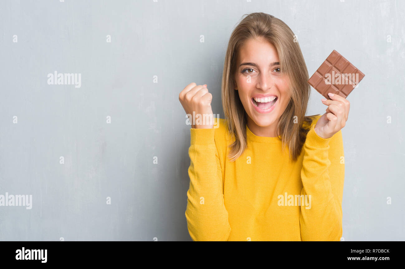 Beautiful young woman over grunge grey wall eating chocolate bar ...