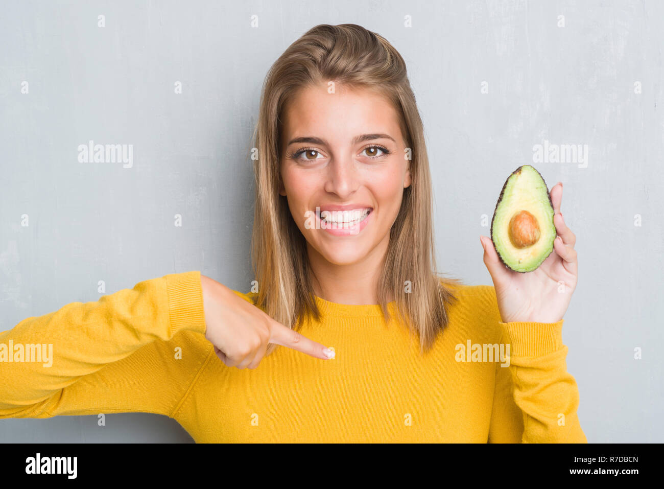 Beautiful young woman over grunge grey wall eating avocado with ...