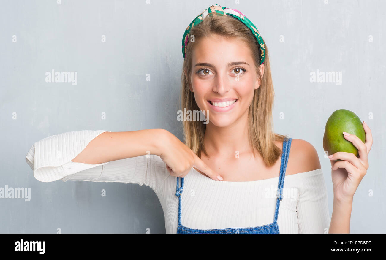 Beautiful young woman over grunge grey wall holding fresh mango with ...