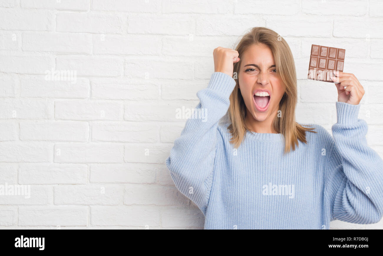 Beautiful young woman over white brick wall eating chocolate bar ...