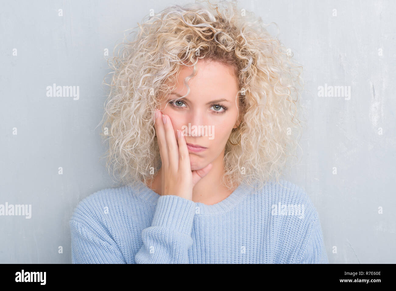 Young blonde woman with curly hair over grunge grey background thinking ...