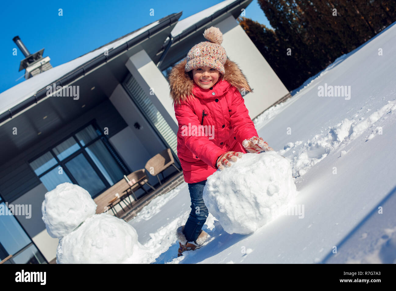 Winter vacation. Girl standing outdoors near house rolling snowball ...