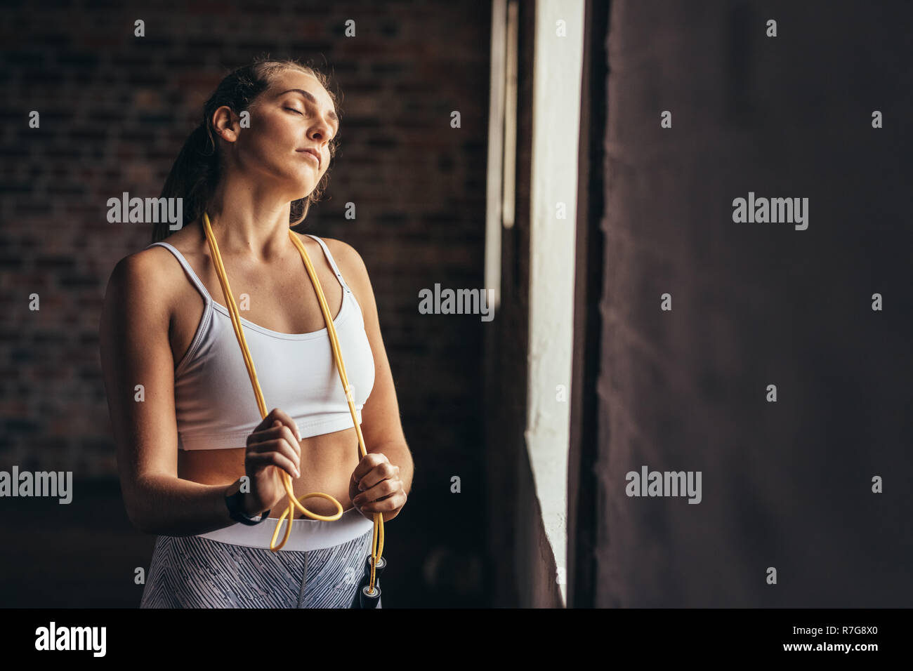 Woman athlete with skipping ropes around her neck standing by gym ...