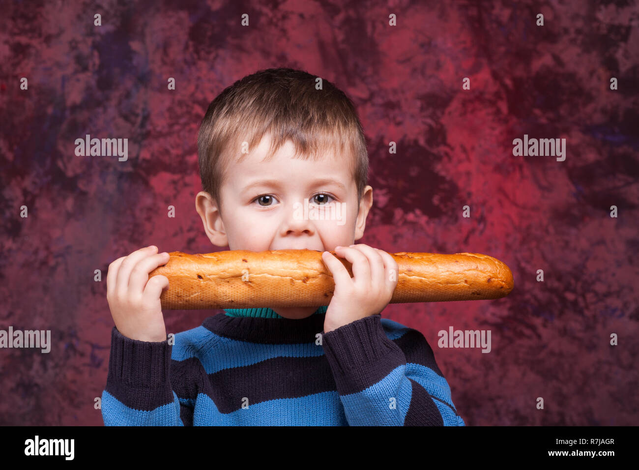 Cute kid holding and biting French bread against dark red background ...