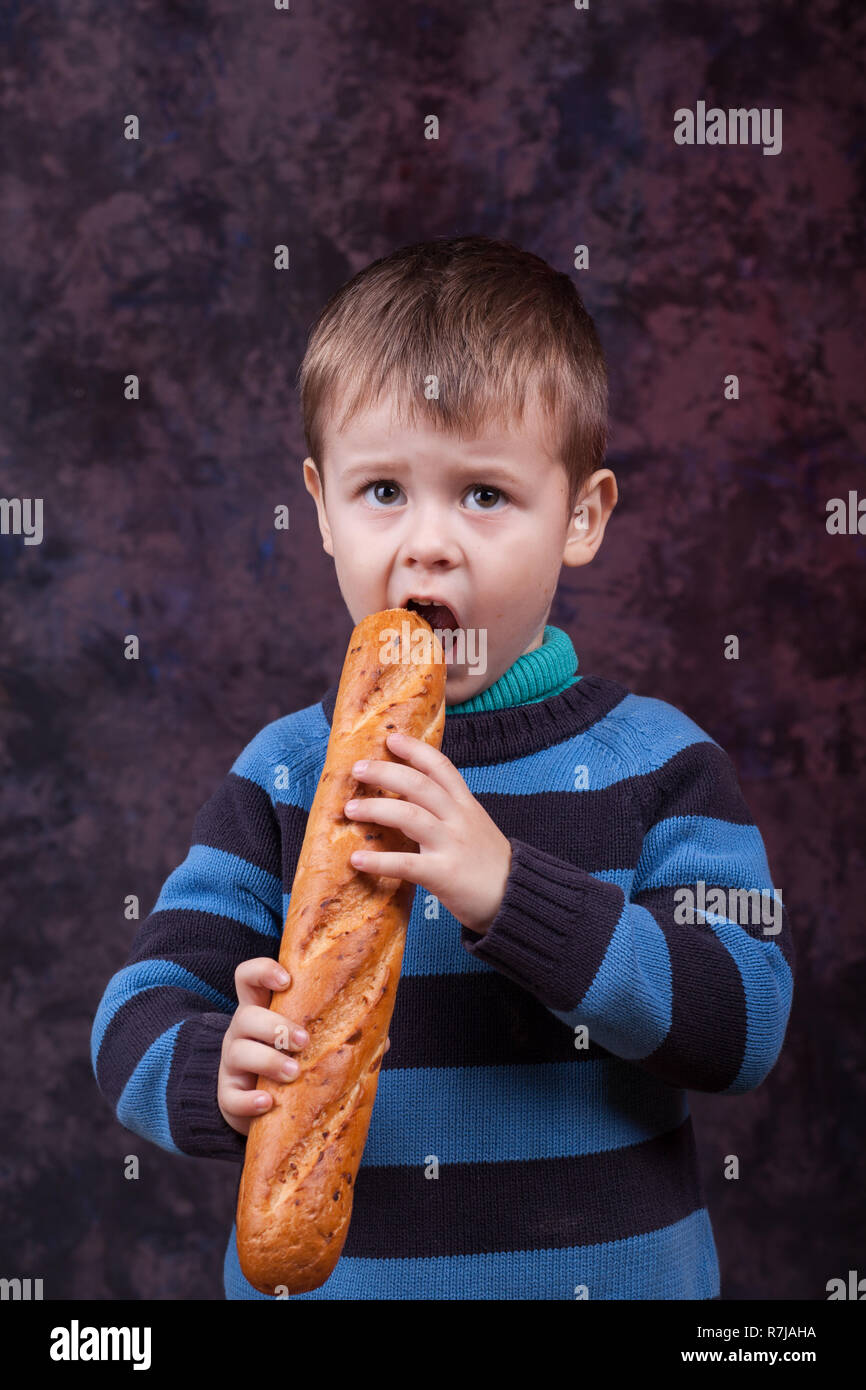 Cute kid holding and biting French bread against dark red background ...