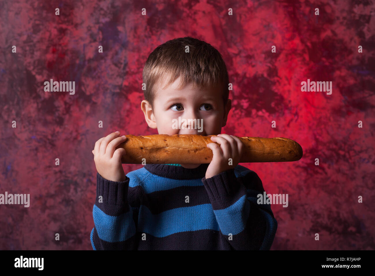 Cute kid holding and biting French bread against dark red background ...