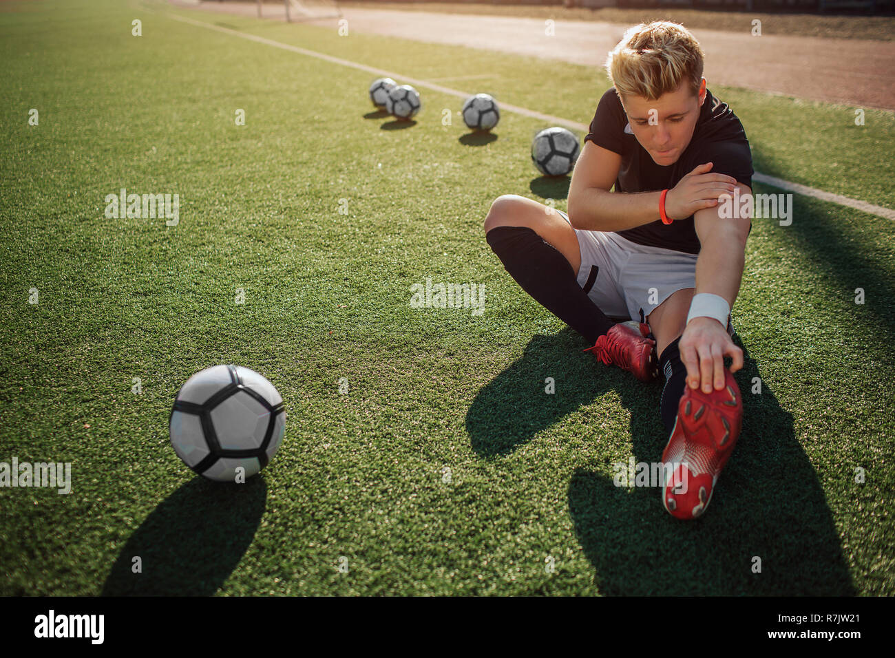 Young blonde football player sit on lawn and stretch. He reaches with ...