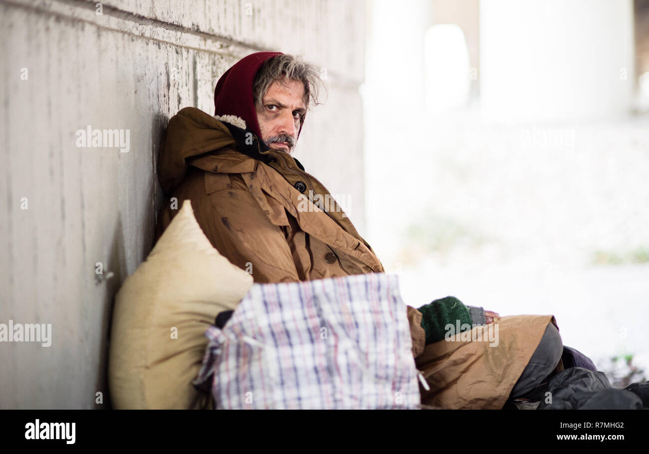 A side view of homeless beggar man sitting outdoors, leaning against a ...