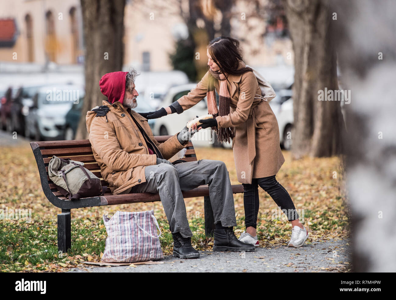 Young Woman Giving Food To Homeless Beggar Man Sitting On A Bench In
