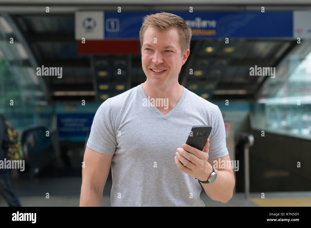 Young happy tourist man thinking while using phone at the train station ...