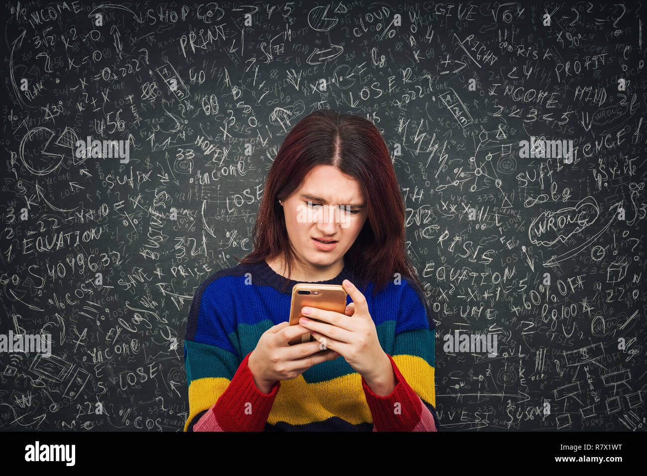 Confused young woman in front of a blackboard looking at her smartphone ...