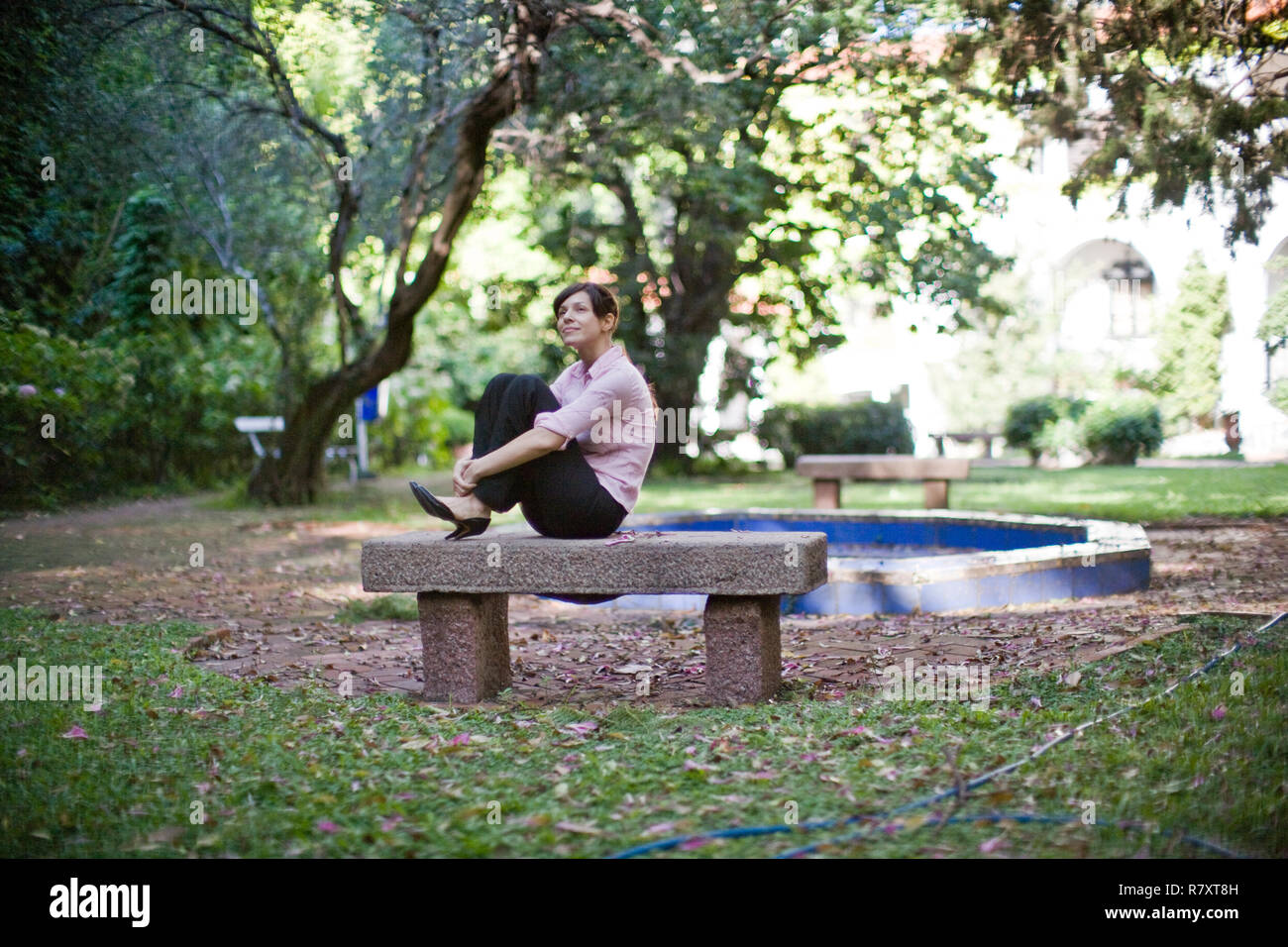 Mid-adult woman sitting on a park bench with her arms around her legs ...