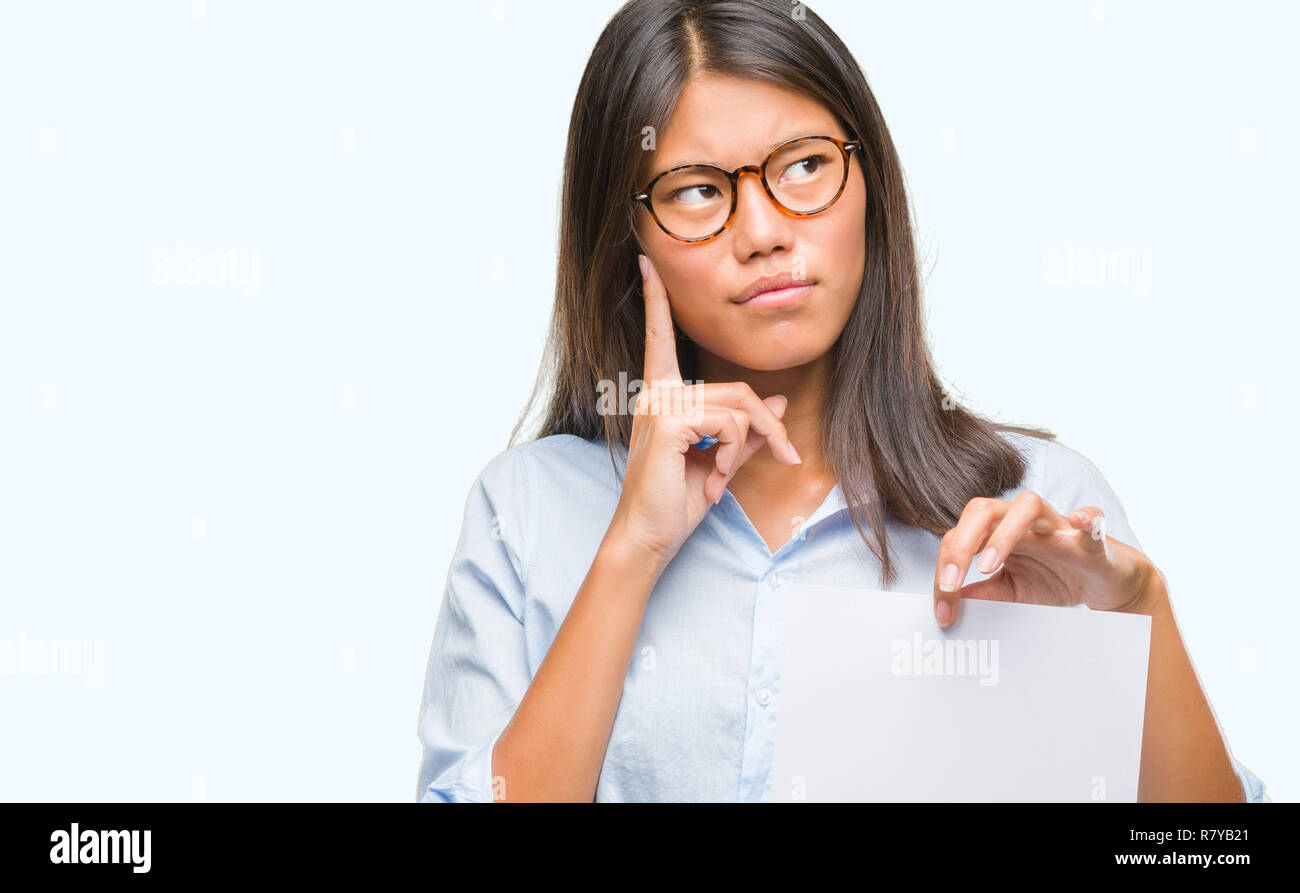 Young asian woman over isolated background holding blank paper serious face thinking about ...