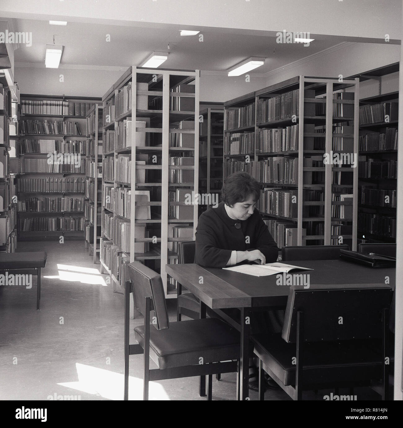 1960s, historical, a young lady sitting alone at a table in a library ...