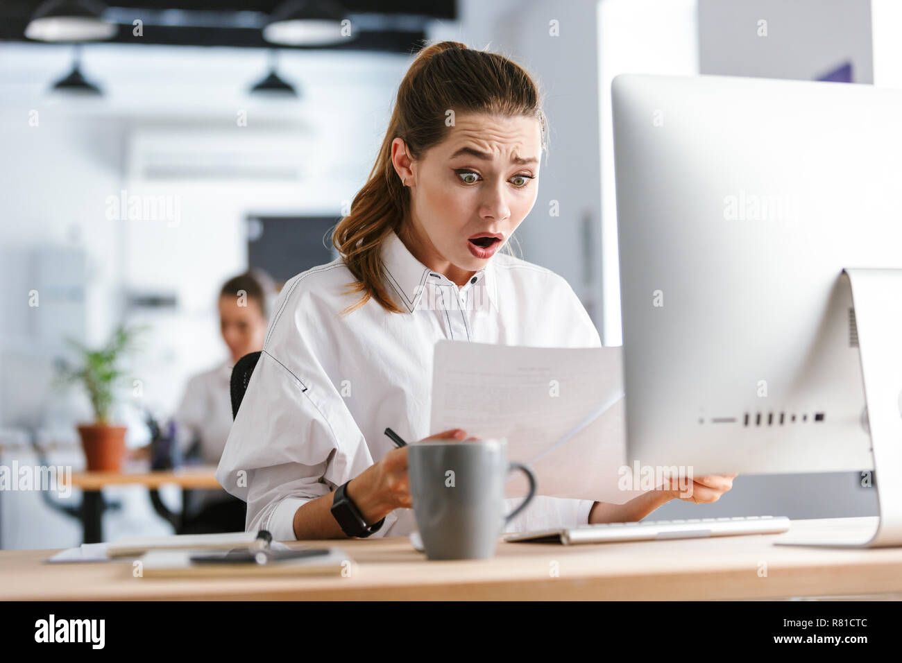 Shocked young woman dressed in shirt sitting at her workplace at the ...