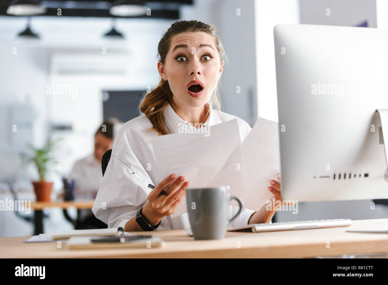 Shocked young woman dressed in shirt sitting at her workplace at the ...
