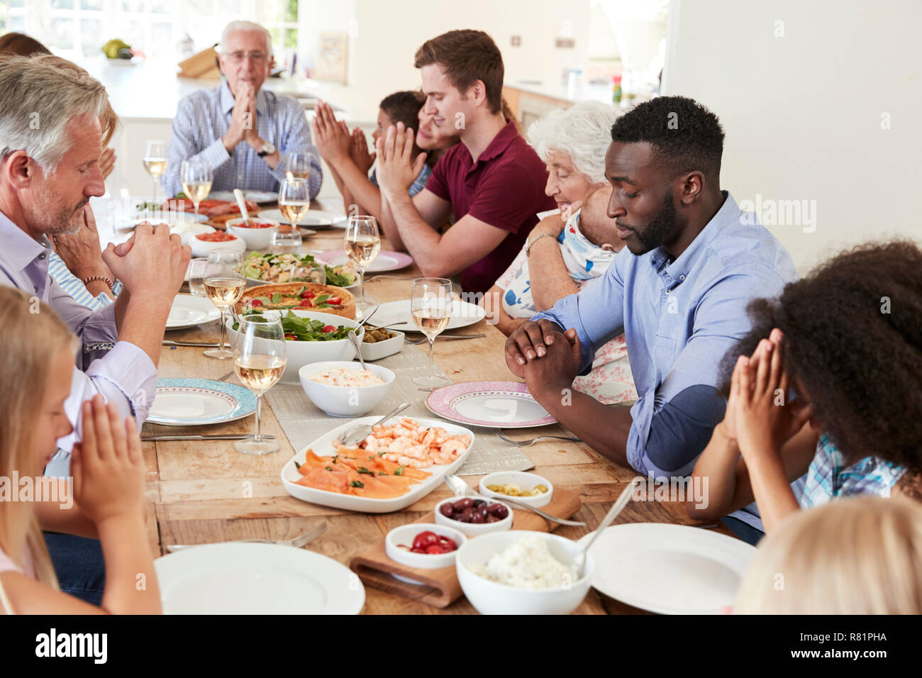 Multi-Generation Family And Friends Around Table Praying Before Meal At ...