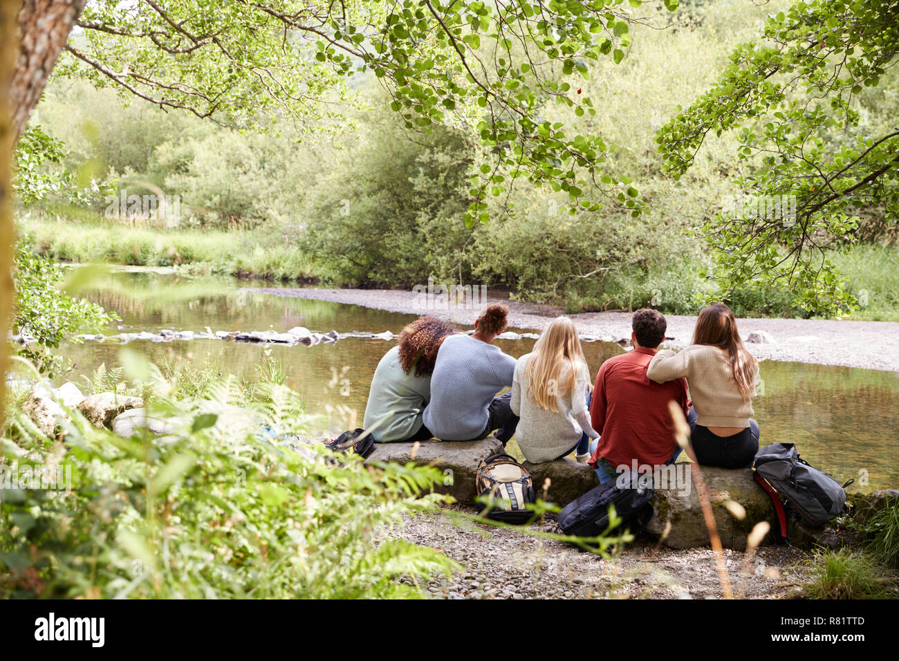 Five young adult friends taking a break sitting on rocks by a stream ...