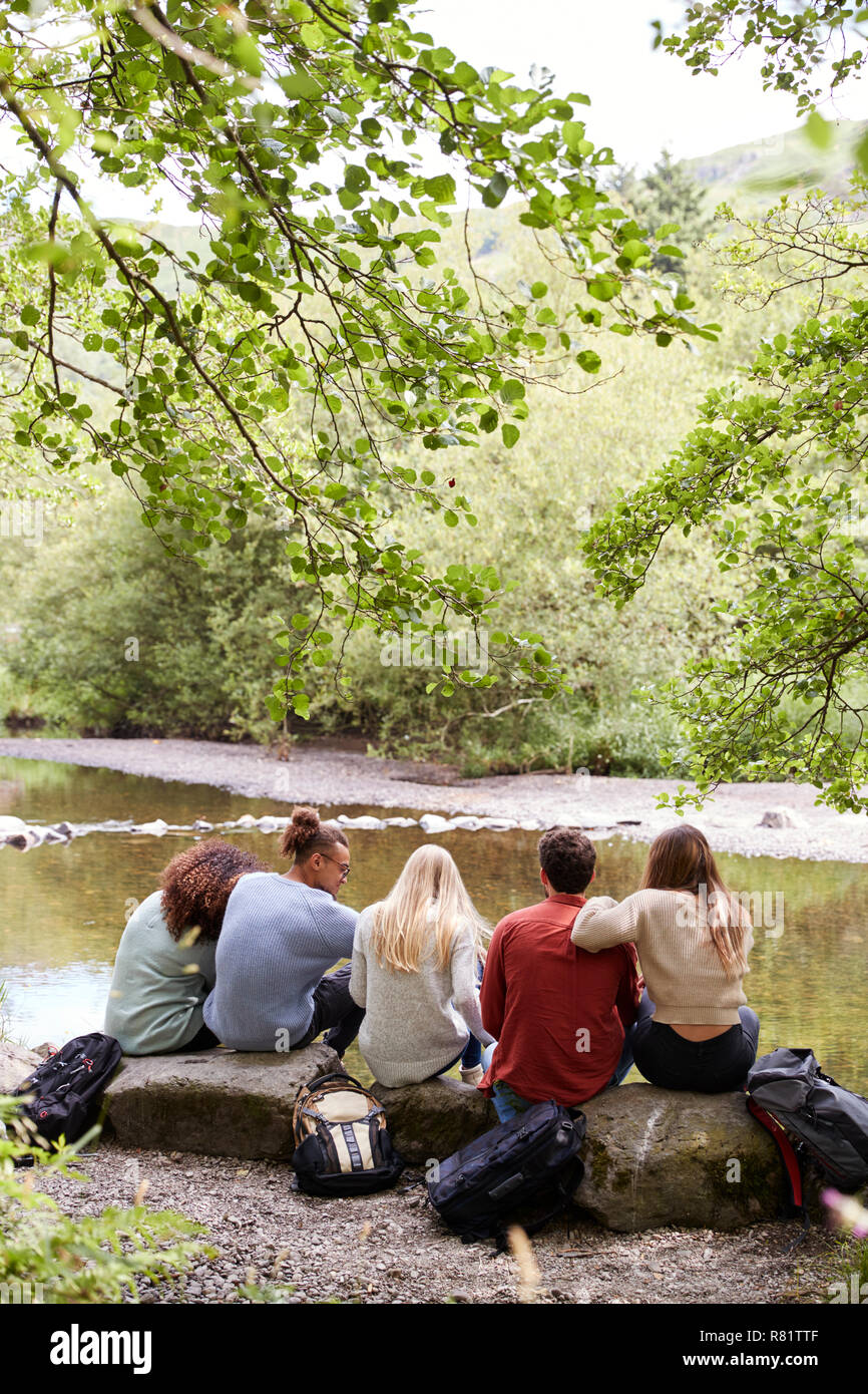 Five young adult friends taking a break sitting on rocks by a stream ...