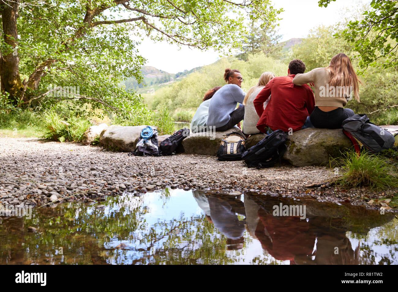 Five young adult friends taking a break sitting on rocks by a stream ...